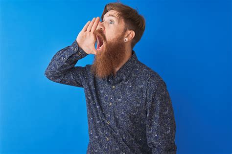 고립 된 파란색 배경 위에 서있는 꽃 여름 셔츠를 입은 젊은 빨간 머리 아일랜드 남자가 입에 손을 들고 큰 소리로 외치고 비명을 지르고 있습니다 통신 개념입니다 감정에 대한