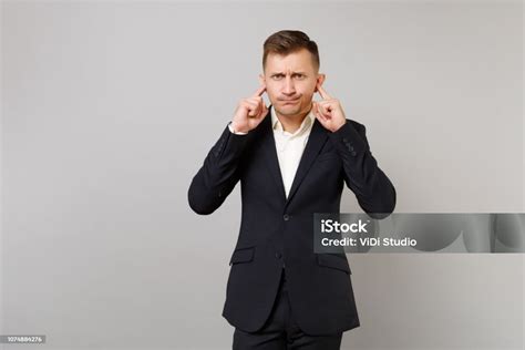 스튜디오에서 회색 벽 배경에 고립 된 손가락으로 귀를 덮고 고전적인 검은 양복에 불만족된 젊은 사업가의 초상화 성과 경력 부 비즈니스 개념입니다 복사 공간을 비웃는 다 검은
