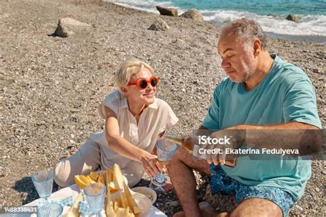 여름 해변 피크닉 해변의 해변에서 신선한 과일 와인 애피타이저로 피크닉을 즐기는 중년 부부 금발 여자를 위해 와인잔에 와인을 붓는 수염 난 남자 2명에 대한 스톡 사진 및