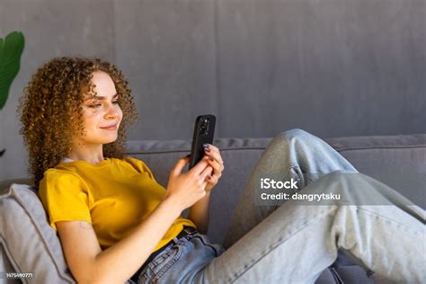 측면보기 젊은 충격을받은 여자는 흰색 탱크 셔츠를 입고 휴대 전화를 사용하여 파란색 소파에 앉아 집에 머물러 평면 휴식 휴식