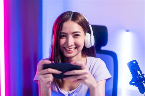 휴대 전화에서 비디오 게임 승리 거실에서 스마트 폰을 들고 의자에 앉아있는 젊은 아시아 예쁜 여자 행복한 여성 프로 스트리머 중국어 착용 헤드폰 네온 라이트와 온라인 게임을