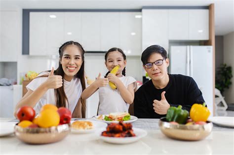 아시아 가정이 함께 식사를 하고 집에서 엄지 손가락을 행복하게 보여주고 행복한 젊은 부모들은 식탁에서 점심을 먹으며 어린 딸과 함께 즐거운 시간을 보내고 있습니다 20 24