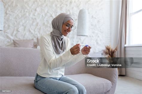 옆에서 사진 히잡을 쓴 젊은 무슬림 여성이 집 소파에 앉아 일회용의 빠른 임신 테스트기를 들고 웃고 있다 긍정적 인 결과에