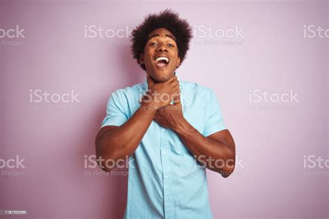 고립 된 분홍색 배경 위에 서있는 파란색 셔츠를 입은 아프리카 머리를 가진 젊은 미국 남자는 고통스러운 목졸림 때문에 외치고 질식합니다 건강 문제 질식 과 자살 개념 건강