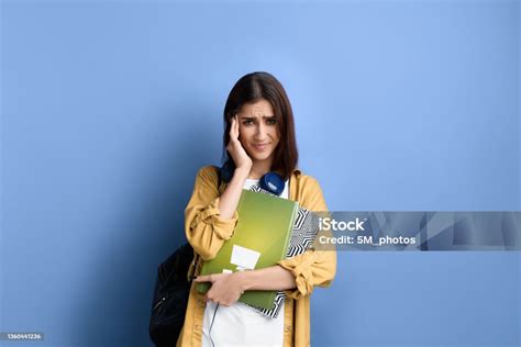 피곤한 학생 소녀는 손가락으로 사원을 찡그린 채 두통이나 편두통이 있고 공부를 다치고 책을 들고 노란색 셔츠 흰색 티셔츠 검은 색 가방 헤드폰을 목에 걸고 있습니다 20