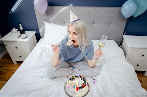 파자마와 파티 모자를 쓴 슬프고 화가 난 여자는 샴페인을 마시고 장식된 침실의 침대에 앉아 생일 케이크를 먹고 있다 혼자 생일을