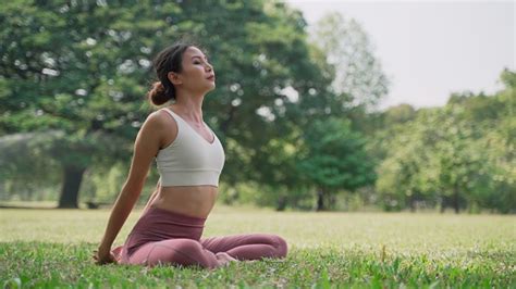 연꽃 자세의 잔디에 앉아 큰 나무 배경을 가진 도시 공원에서 손을 들고 있는 아시아 청년 여성 화창한 날에 는 야외에서 요가를 연습하는 여성의 백미러 30 34세에 대한