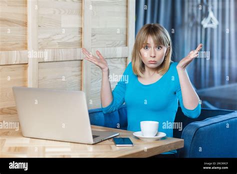 Confused Uncertain Young Woman With Blonde Hair In Blue Shirt Sitting
