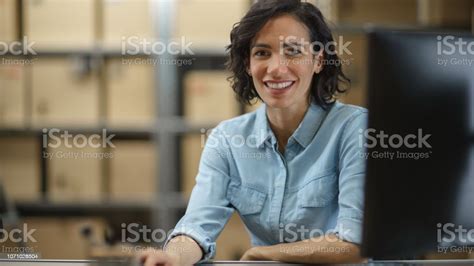 그녀의 책상에 앉아 작업 컴퓨터 및 카메라에 미소를 작동 하는 여성 재고 관리자 배경 창 고 저장실 선반에 골 판지 상자의 전체 패키지 배송 준비 여자에 대한 스톡 사진 및