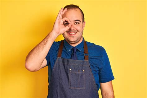 고립된 노란색 배경 위에 서있는 앞치마를 입은 젊은 상점 주인 남자는 행복한 얼굴로 손가락을 통해 보는 손 미소와 함께 확인 제스처를하고 있습니다 소매 관련 직업에 대한