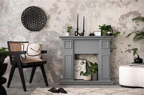 회색 벽난로 흰색 Pouf 세련된 등나무 안락 의자 화분에 식물 무늬가있는 카펫 및 개인 액세서리가있는 거실 인테리어의 창조적 인 구성 가정 장식 템플렛 벽난로에 대한 스톡