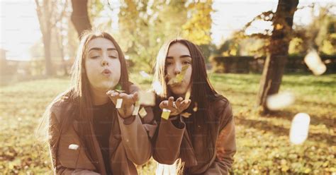 젊은 갈색 머리 쌍둥이 자매는 서로 가까이 서서 색종이를 카메라에 불고 그 중 일부를 손에 들고 흐릿한 배경에 가을 햇볕이 잘 드는 공원에서 캐주얼 코트를 입고 있습니다