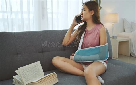 Young Asian Woman Wearing Soft Splint Due To Sore Arm Using Phone On Sofa At Home Stock Image