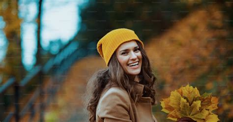 안녕하세요 가을입니다 갈색 코트와 노란 모자를 쓴 행복한 세련된 여자는 가을에 도시 공원에 밖에 가을 노란 잎을 가지고 있다