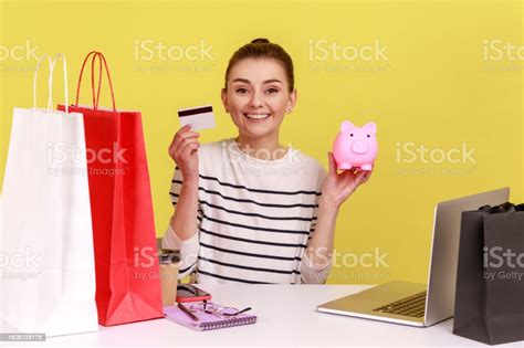 쇼핑백을 들고 직장에 앉아 있는 여자는 돼지 저금통과 신용카드를 카메라에 보여준다 25 29세에 대한 스톡 사진 및 기타 이미지 25 29세 가계 계산함 Istock
