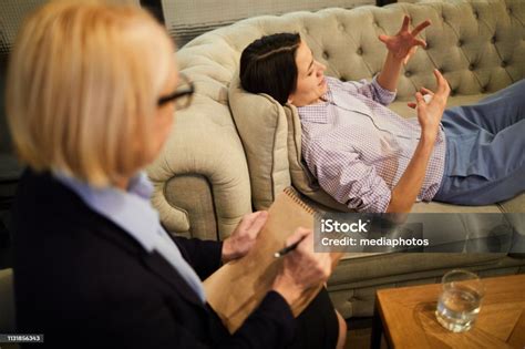 소파와 몸짓 손에 누워 캐주얼 셔츠에 화가 젊은 여자 치료사에 그녀의 감정을 설명 하는 동안 여자 정신과 의사 만들기 노트 스케치 Mental Wellbeing에 대한 스톡