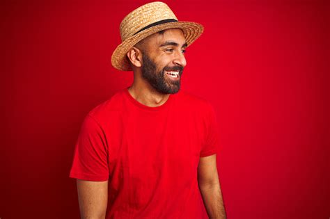 티셔츠와 모자를 쓰고 있는 젊은 잘생긴 인도 남자가 고립된 빨간 배경 위에 얼굴을 미소로 옆으로 바라보며 자연스러운 표정을 짓고