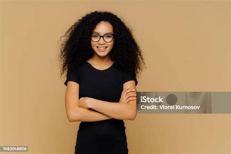 아프로 헤어 스타일로 슬림 만족 여자 검은 캐주얼 옷을 입고 광학 안경 자신감 표정을 가지고 베이지 색 배경에 고립 된 대담을 듣고 귀여운 십대 소녀는 인생을 즐긴다 T