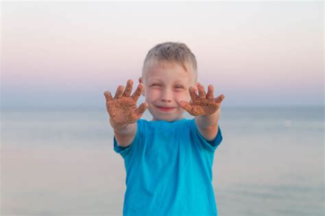 아름다운 소년은 저녁에 바다의 배경에 파란색 티셔츠에 유럽을 금발 아이는 바다 모래에 더럽혀진 손을 보여줍니다 아기는 미소를 지었다 바다 또는 바다에서 아이들을위한 멋진