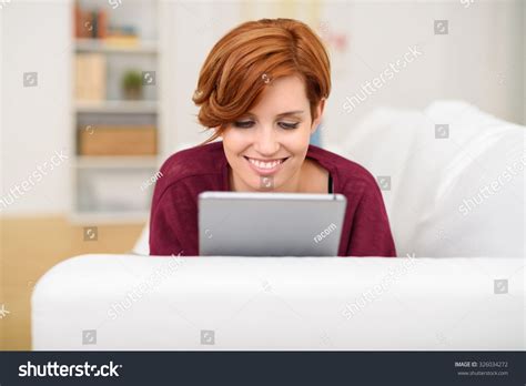 Attractive Young Redhead Woman Relaxing On A Sofa In The Living Room Using A Tablet To Browse