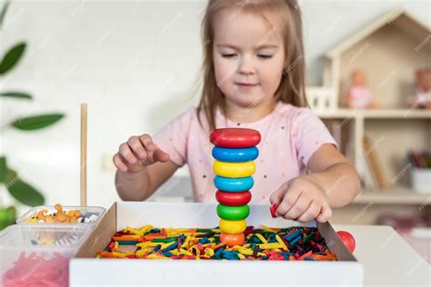 나무 피라미드와 색색의 파스타를 가지고 노는 어린 소녀 어린이를 위한 감각 놀이 및 학습 색 활동 미세 운동 기술을 위한 몬테소리 게임 감각 상자 교육 장난감 프리미엄 사진