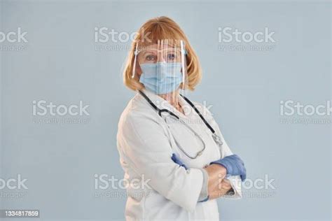 흰색 코트를 입은 수석 여성 의사가 파란색 배경에 대해 실내에 서 있습니다 65 69세에 대한 스톡 사진 및 기타 이미지