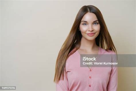 매력적인 유럽 여성의 수평 초상화는 카메라에서 부드럽게 미소 긴 직선 머리를 가지고 캐주얼 장미 스웨터를 입고 야외 산책을하려고 베이지 색 스튜디오 배경에 대해 서있다 25