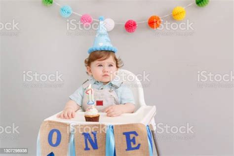 집에서 첫 번째 생일을 축하하는 파란색 모자를 쓴 귀여운 사랑스러운 백인 아기 소년 밝은 촛불이달린 컵케이크 디저트를 바라보며 높은 의자에 앉아 있는 아이 유아 생일 컨셉
