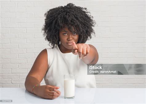 카메라 하 고 당신에 게 손 기호 긍정적이 고 자신감 제스처 앞에서 손가락으로 가리키는 우유 한 잔을 마시는 테이블에 앉아 젊은 아프리카계 미국인 여자 가리키기에 대한 스톡