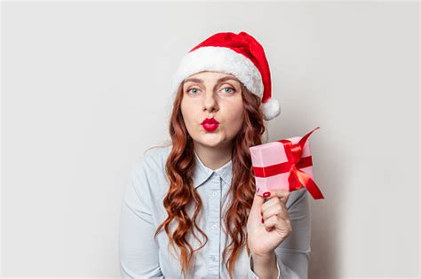 곱슬 머리와 범본과 빨간 모자와 산타 클로스 소녀는 빨간색 새틴 리본 활과 선물 상자를 보유하고 회색 벽의 배경에 카메라를보고