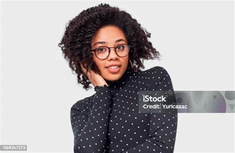 예쁜 꿈같은 검은 피부젊은 여성은 검은 색으로 옷을 입고 흰색 점 블라우스를 입고 머리에 손을 잡고 행복을 느낍니다 흰 벽에 포즈를 취하는 둥근 안경을 쓰고 활짝 웃고 있는
