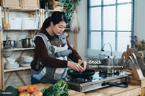 캐리어에 아기를 가진 아시아 주부는 밝은 가정 부엌에서 아침 식사를하는 동안 프라이팬에 계란을 깨고있다 요리하기 음식 준비에 대한 스톡 사진 및 기타 이미지 Istock