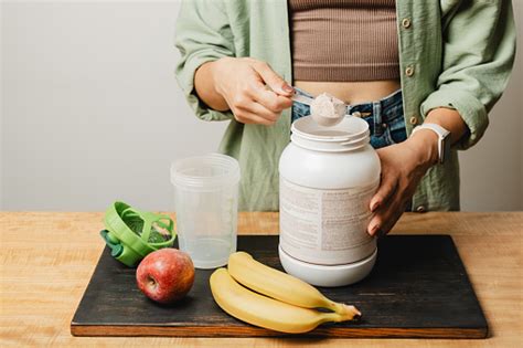청바지와 셔츠를 입은 여자는 셰이커 바나나 사과 과일이 있는 나무 테이블 위에 있는 플라스틱 병 위에 유청 단백질 분말의 일부가