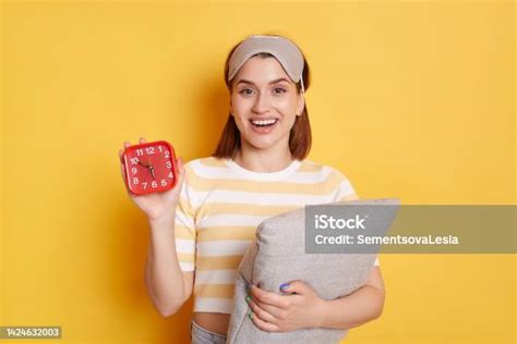 회색 베개와 빨간 알람 시계를 들고 흥분한 카메라를보고 고립 된 노란색 배경을 포즈를 취하는 흥분한 행복한 만족 어두운 머리 여자의 사진 벽 시계에 대한 스톡 사진 및 기타