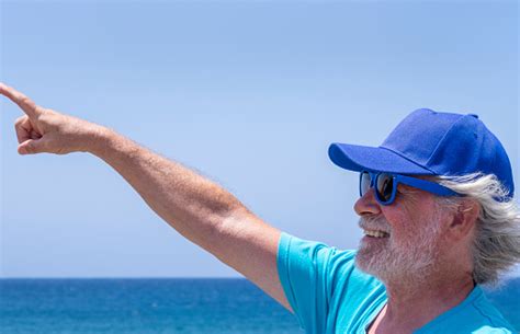 하얀 머리를 웃고 해변에 서있는 노인 수염 남자의 초상화는 먼 지점 푸른 바다와 배경의 하늘을 가리키는 해변에 서 활동적인 행복한 노인의 개념 건강한 생활방식에 대한 스톡