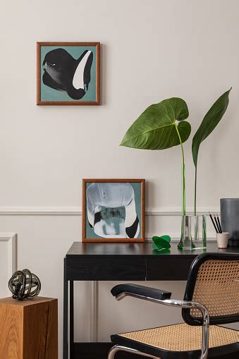 모형 포스터 프레임 검은색 책상 등나무 의자 나뭇잎이 있는 세련된 꽃병 다채로운 조각품 벽에 치장 벽토 및 개인 액세서리가 있는 작업장 인테리어의 현대적인 구성 홈 데코
