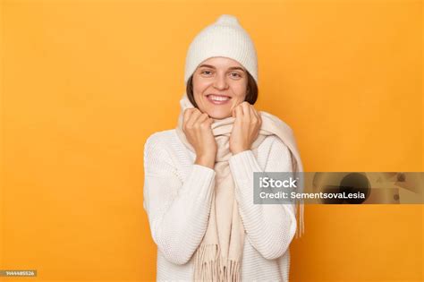 흰색 점퍼 모자 스카프를 착용하고 웃고 있는 긍정적인 즐거운 여성의 이미지는 옷깃에 손을 얹고 이빨 미소로 카메라를 바라보며 노란 벽에 포즈를 취합니다 30 34세에 대한