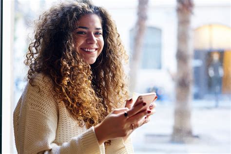 흐리게 프로 모션 배경 블로깅 소셜 네트워크에 대 한 Wifi에 연결 하는 전화를 들고 카페에서 시간을 보내는 카메라 보고 곱슬 머리를 가진 쾌활 한 아름 다운 젊은 여자의