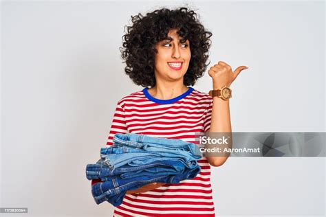 젊은 아랍 상점 주인 여자 와 곱슬 머리 들고 청바지 위에 고립 된 흰색 배경 가리키는 고 행복 한 얼굴 미소와 측면에 엄지 손가락으로 표시 더미에 대한 스톡 사진 및 기타