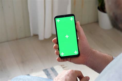 스마트폰 녹색 모형 화면을 손에 들고 집에 앉아 있는 청년 크로마 키 휴대 전화를 사용하는 남성 수직 모드 터치 디스플레이 스 와이프 탭 인터넷 소셜 미디어 서핑 스크롤에