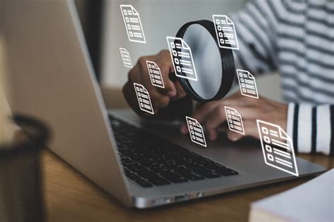 책상 위에 노트북을 놓고 책상에 앉아 있는 한 청년이 돋보기를 사용하여 문서 파일 아이콘을 보고 있습니다 돋보기 광학 기기에