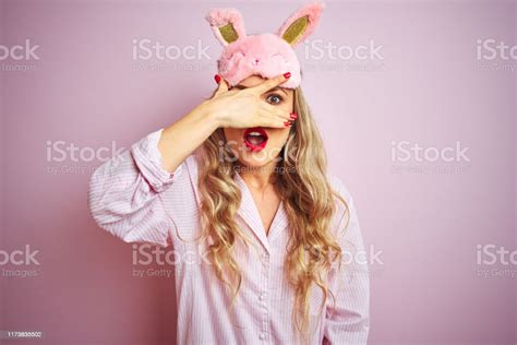 분홍색 고립된 배경 위에 잠옷과 수면 마스크를 쓰고 있는 젊은 아름다운 여성은 얼굴과 눈을 손으로 덮고 부끄러운 표정으로 손가락을 들여다보고 있습니다 가리기에 대한 스톡