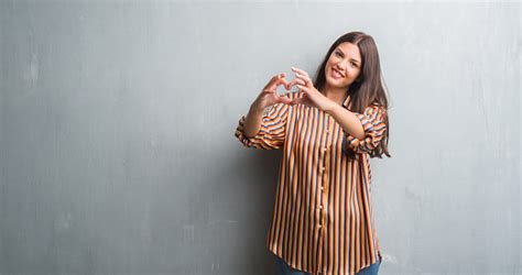 젊은 갈색 머리 여자 마음 기호와 손으로 모양을 보여주는 사랑에 웃 고 그런 지 회색 벽에 낭만적인 개념입니다 갈색 머리에 대한 스톡 사진 및 기타 이미지 Istock