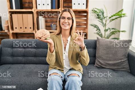 치료 사무실에서 일하는 젊은 금발 여자는 손가락으로 확인 기호를 하고 있는 뇌를 들고 있으며 친절한 몸짓으로 훌륭한 상징을 웃고