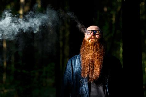Vape 수염 남자 안경에 매우 긴 수염을 가진 성인 흰색 짧은 머리 남자는 어두운 숲에서 외부 전자 담배를 연기 나쁜 습관 건강하지 못한 생활방식에 대한 스톡 사진 및