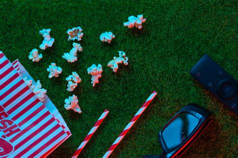 Tv 리모컨으로 영화를 보기 위한 3d 안경이 있는 녹색 잔디밭에 소다를 위한 팝콘 욕조를 포장합니다 빨간색과 파란색의 창조적 인 빛 자연에 대한 영화를 보는 잔디 공원에서