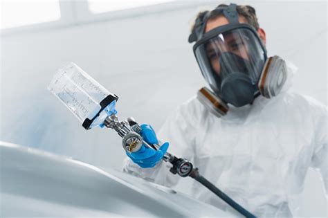 에어브러시 건을 가진 남자가 자동차 범퍼의 표면을 변기합니다 가면을 쓴 자동 화가와 보호복을 입은 자동차 화가가 차고의 자동차 페인팅 챔버에서 작동합니다 수리공에 대한 스톡