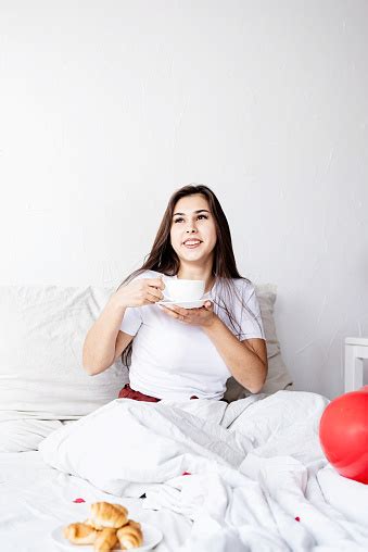 젊은 갈색 머리 여자 앉아 침대에 깨어 있는 빨간 마음 모양풍선 과 장식 마시는 커피 먹는 크루아상 T 셔츠에 대한 스톡 사진