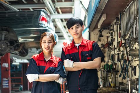 아시아 자동차 엔지니어 사람들은 정비사 차고에서 헬멧 작업을 착용합니다제복을 입은 젊은 자동차 정비사는 카메라를 보고 웃으며 차를 검사하고 있습니다 수리에 대한 스톡 사진