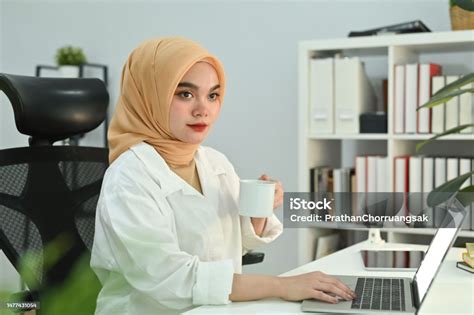 화려한 무슬림 사업가는 히잡을 쓰고 뜨거운 커피를 마시고 사무실에서 노트북 컴퓨터로 작업합니다 25 29세에 대한 스톡 사진 및 기타 이미지 Istock